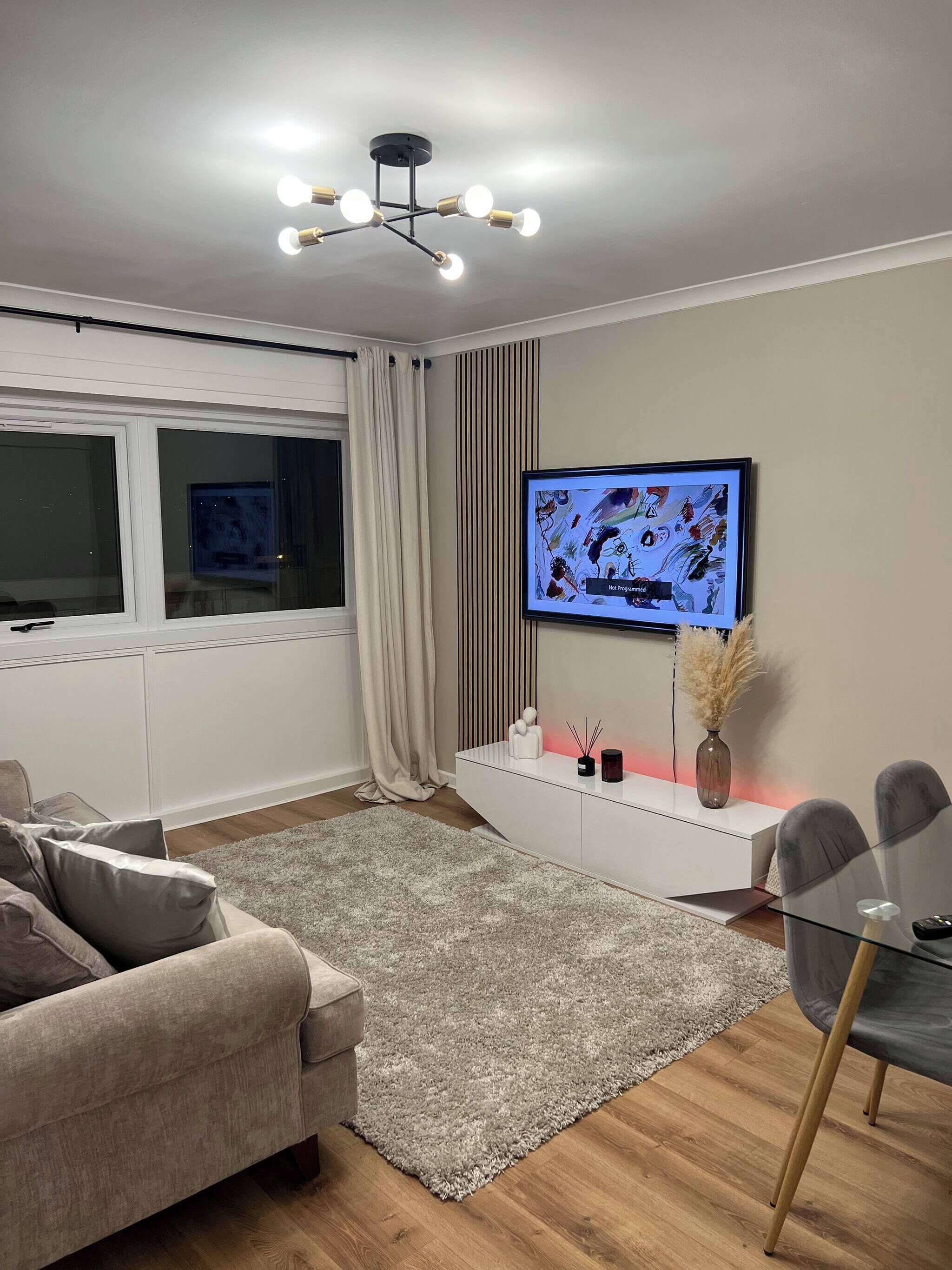 Uma sala de estar moderna com um sofá cinza, uma mesa de centro de vidro, uma TV fixada na parede e um lustre.