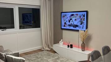 Uma sala de estar moderna com um sofá cinza, uma mesa de centro de vidro, uma TV fixada na parede e um lustre.