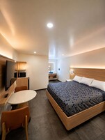 Quarto de hotel moderno com cama, escrivaninha e cadeira, televisão e banheiro ao fundo.