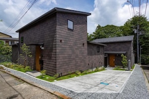 Uma casa moderna de dois andares com fachada escura, uma janela pequena no andar superior e uma entrada frontal.