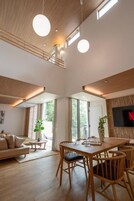 Uma sala de estar moderna com uma mesa de jantar de madeira, cadeiras e um sofá. Há uma TV fixada na parede e uma planta na mesa.