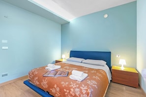 Quarto com uma cama, duas mesas de cabeceira com luminárias, cabeceira azul e colcha florida.