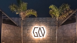Entrada de um edifício moderno com um logotipo geométrico na parede, piso de azulejos e duas palmeiras.