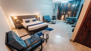 Quarto de hotel moderno com uma cama grande, um espaço de estar com sofá e poltrona, uma mesa redonda pequena e vista para a cidade através de grandes janelas.