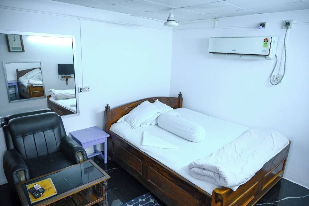 Quarto de hotel com uma cama de madeira, uma mesa pequena com uma cadeira, um espelho, ar condicionado fixado na parede e um relógio.