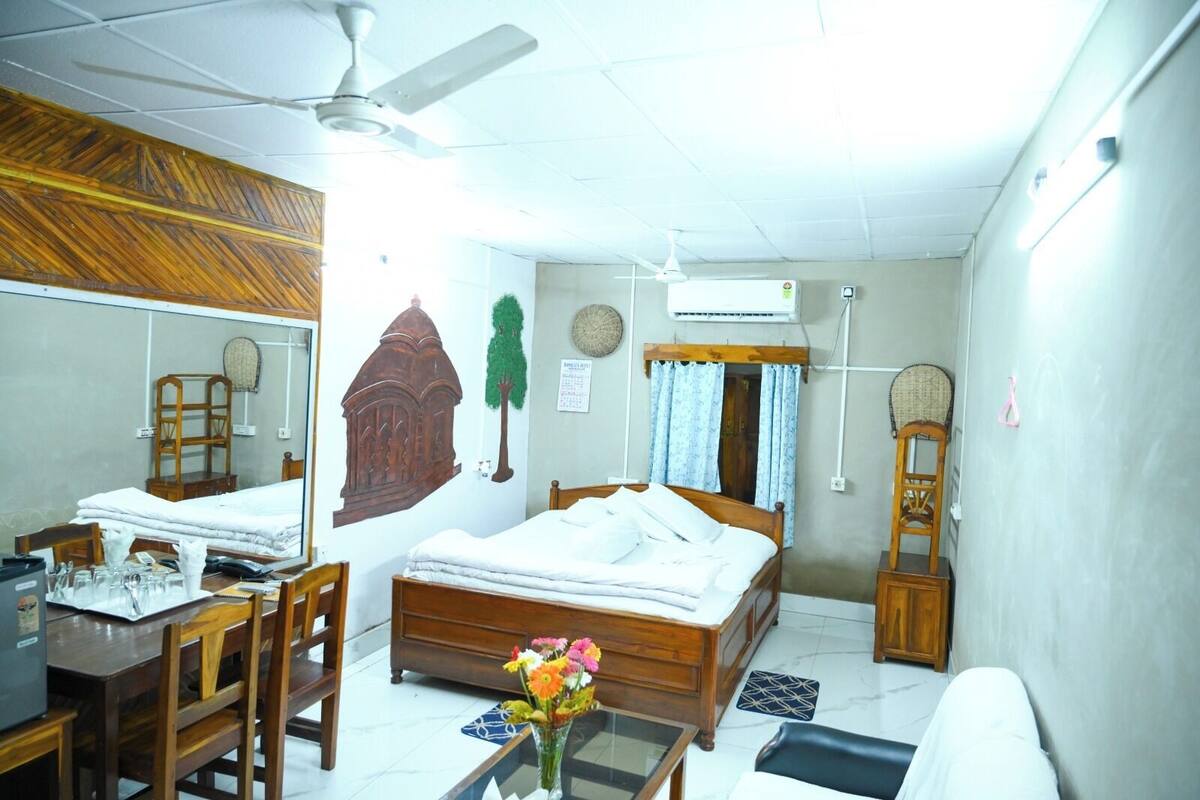 Quarto de hotel com duas camas, uma escrivaninha, uma cadeira, uma mesa com um vaso de flores, um ventilador de teto, um ar condicionado e um ventilador fixado na parede.