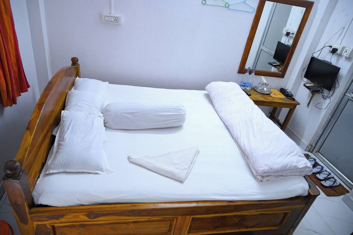 Cama bem arrumada com lençóis brancos, cabeceira de madeira, mesa de cabeceira com abajur e espelho acima.