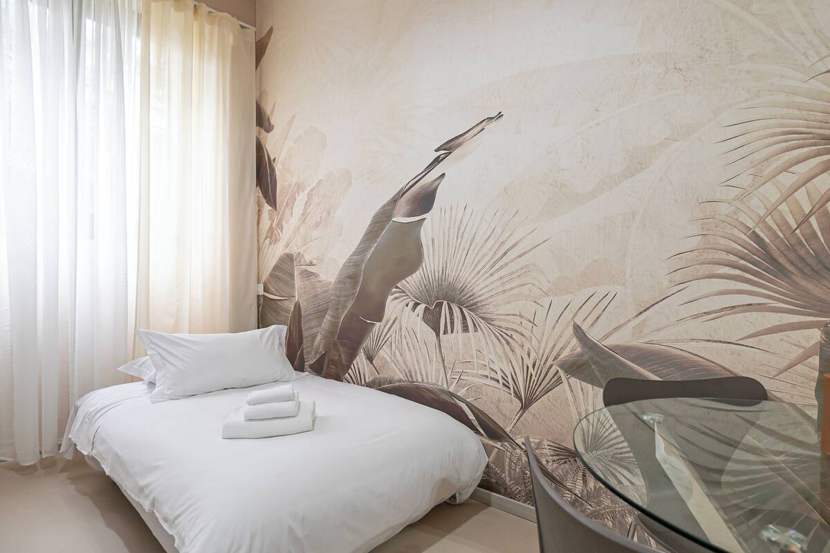 Quarto de hotel com uma cama, uma mesa de vidro e um mural de plantas tropicais.