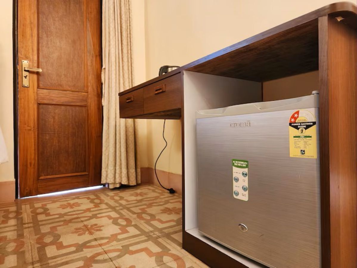 Quarto com porta de madeira, escrivaninha com gavetas e refrigerador metálico.