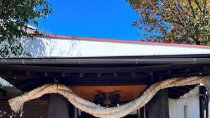 Um edifício japonês tradicional com fachada de madeira, entrada coberta de palha e um caminho de pedra que leva até a entrada.
