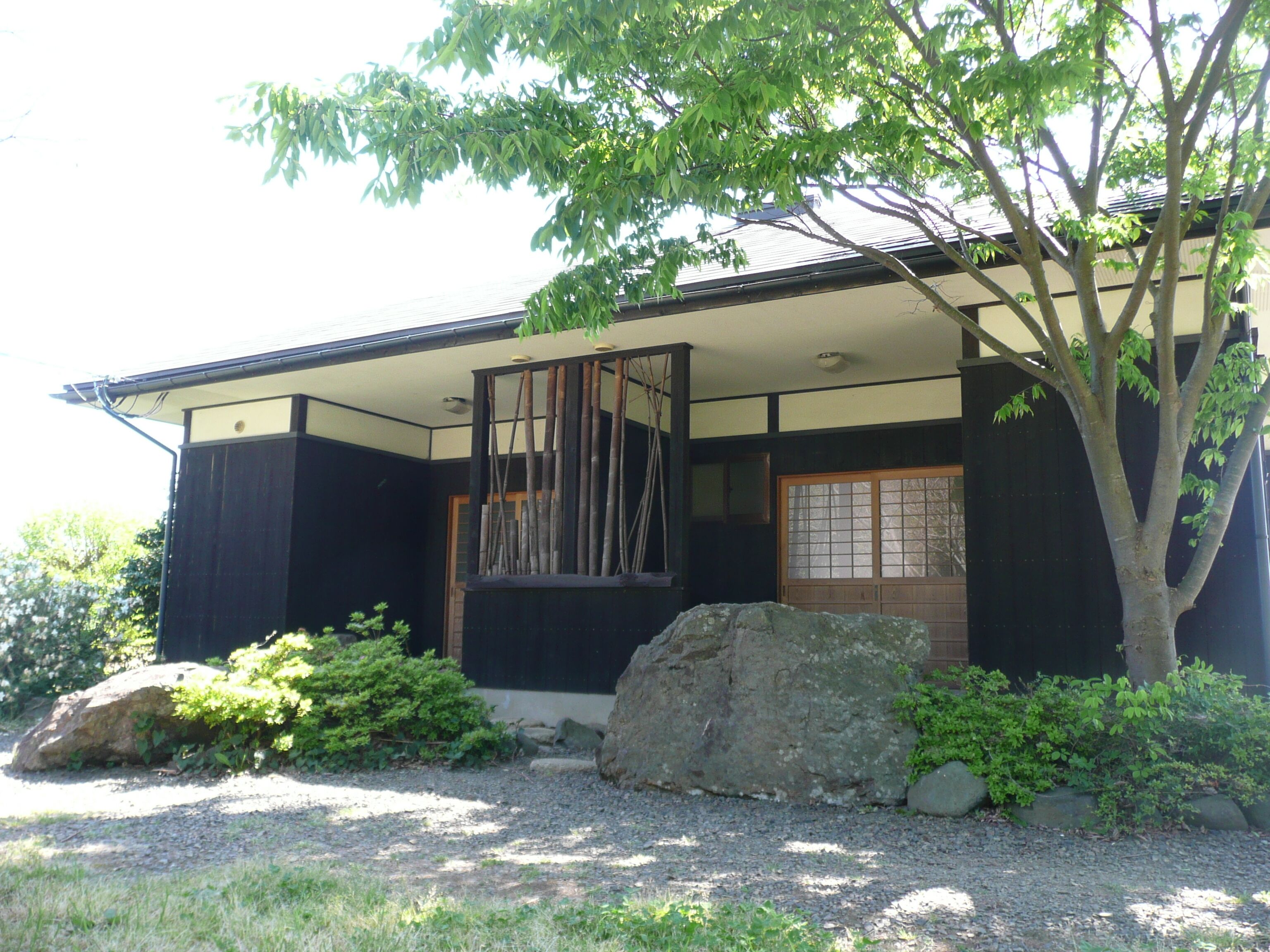 Uma casa japonesa tradicional com fachada de madeira, um amplo jardim de pedras e uma árvore no quintal.