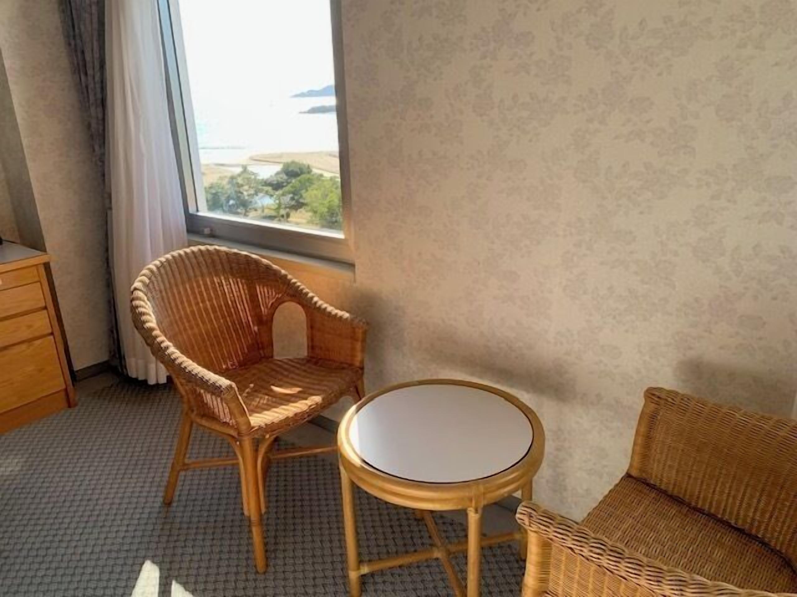 Quarto com uma cadeira de vime, uma pequena mesa redonda e vista para o exterior pela janela.