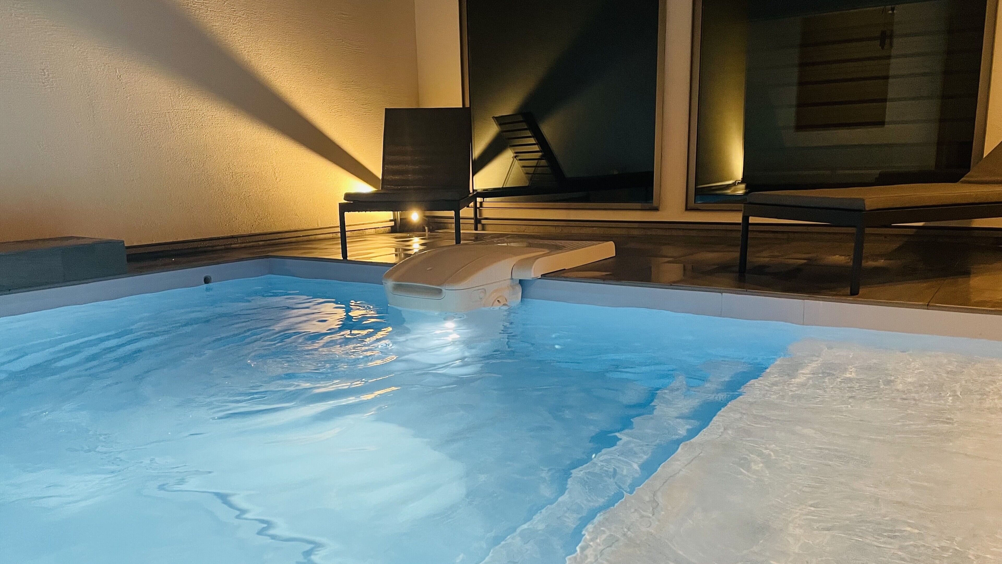 Piscina interior com superfície de água límpida, um banco embutido e vista para uma escada e uma janela ao fundo.