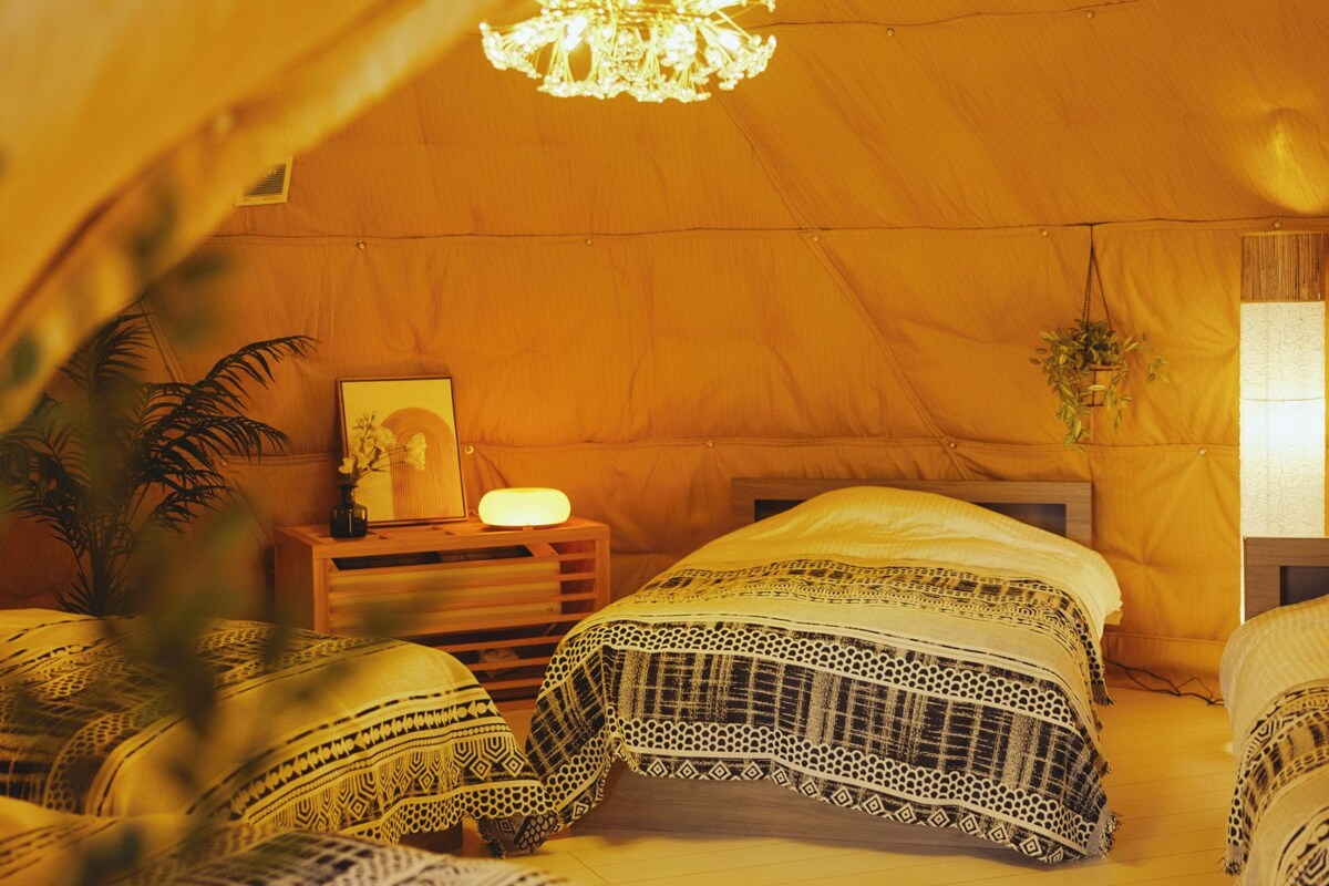 Uma tenda acolhedora com duas camas, um candeeiro pendente, uma mesa pequena com uma fotografia e uma planta suspensa.