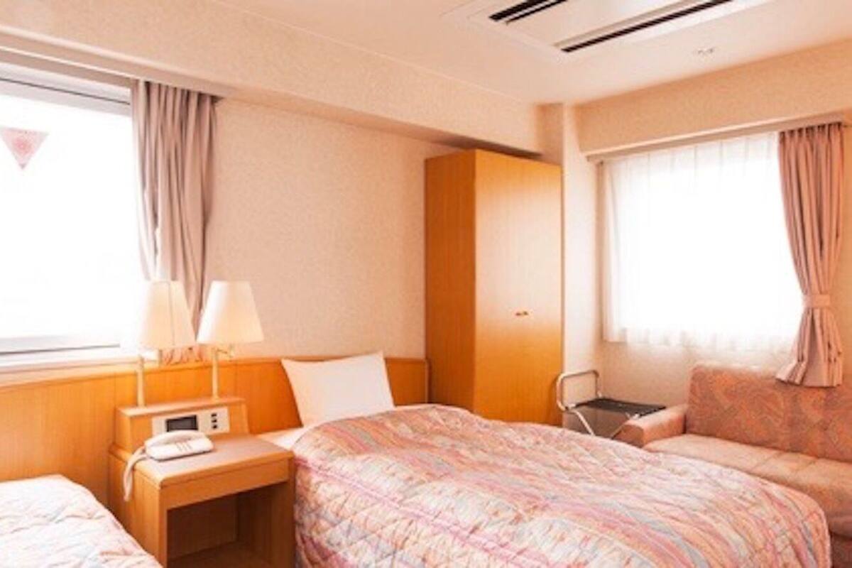 Quarto de hotel com duas camas, uma mesa de cabeceira com abajur, uma mesinha com telefone e uma janela com cortinas.
