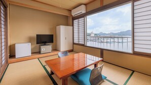 Quarto com piso de tatame, uma mesa de madeira, televisão, mini geladeira e vista para um porto com barcos e montanhas.
