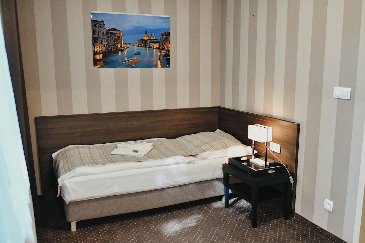 Quarto de hotel com cama, mesa de cabeceira com abajur, quadro com vista urbana e parede listrada.