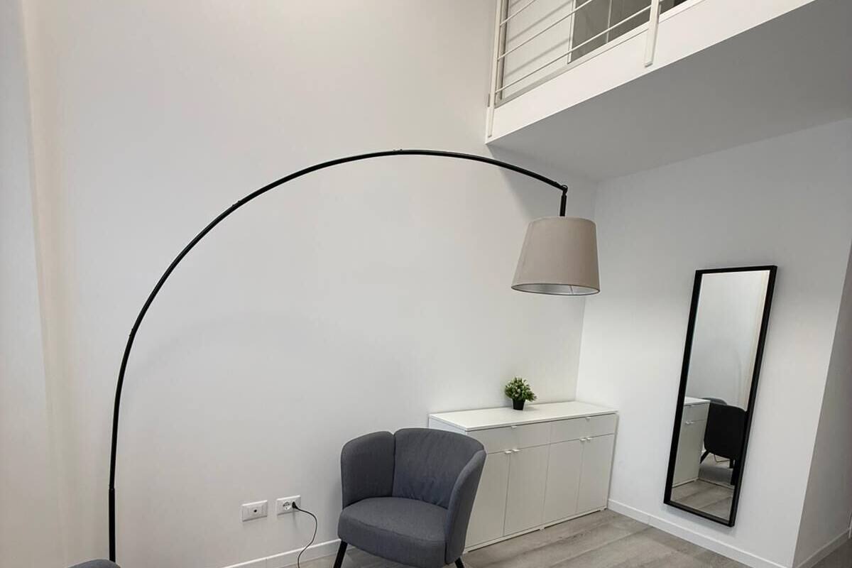 Quarto moderno com sofá cinza, uma escrivaninha com luminária, espelho e janela com persianas.