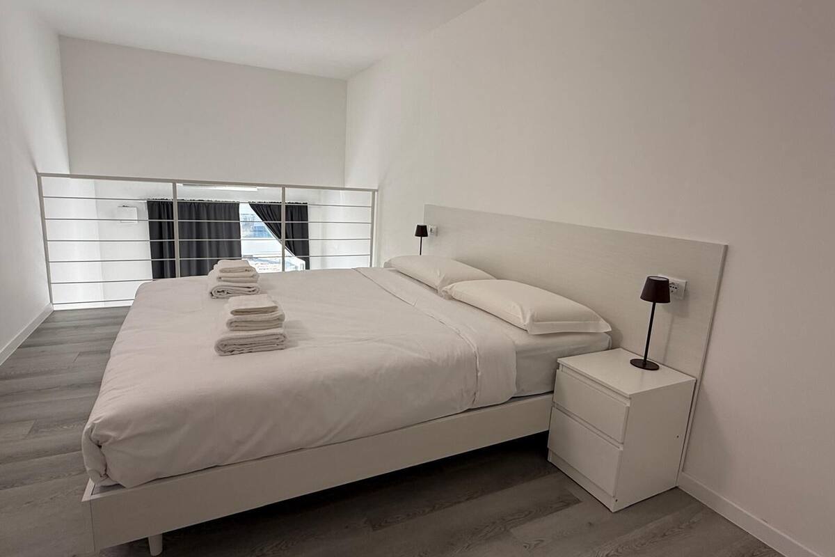 Quarto moderno com cama branca, mesa de cabeceira e vista para uma varanda.