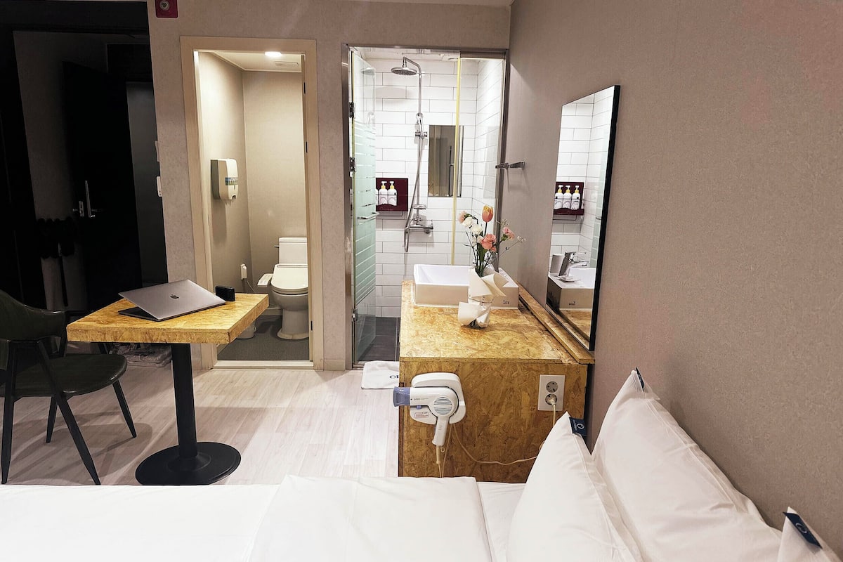 Quarto de hotel com cama, escrivaninha com laptop, cadeira, banheiro com chuveiro, pia e espelho.