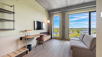 Quarto moderno com sofá, escrivaninha, cadeira, TV e amplas janelas com vista para a cidade.
