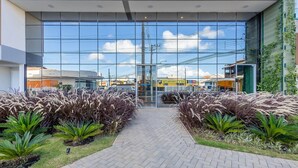 Um edifício moderno com fachada de vidro, entrada pavimentada e vegetação ao redor.