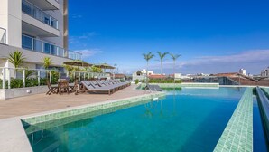Uma piscina no terraço com espreguiçadeiras e guarda-sóis, cercada por vegetação e vista para a cidade.