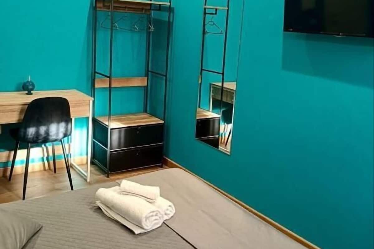Quarto com paredes verde-água, cama com roupa de cama cinza, escrivaninha com cadeira, espelho e televisão na parede.