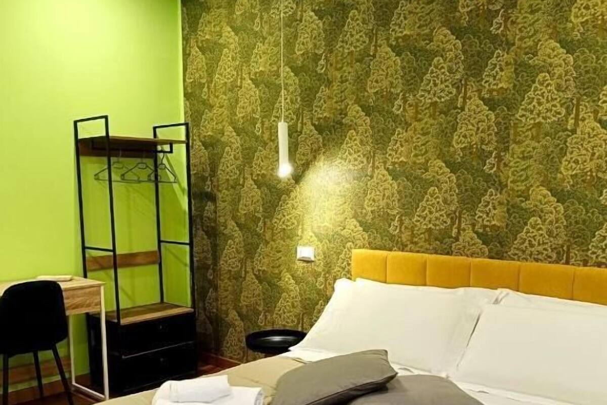 Cama bem arrumada com almofadas e toalhas enroladas, uma escrivaninha com cadeira e parede com papel de parede estampado.