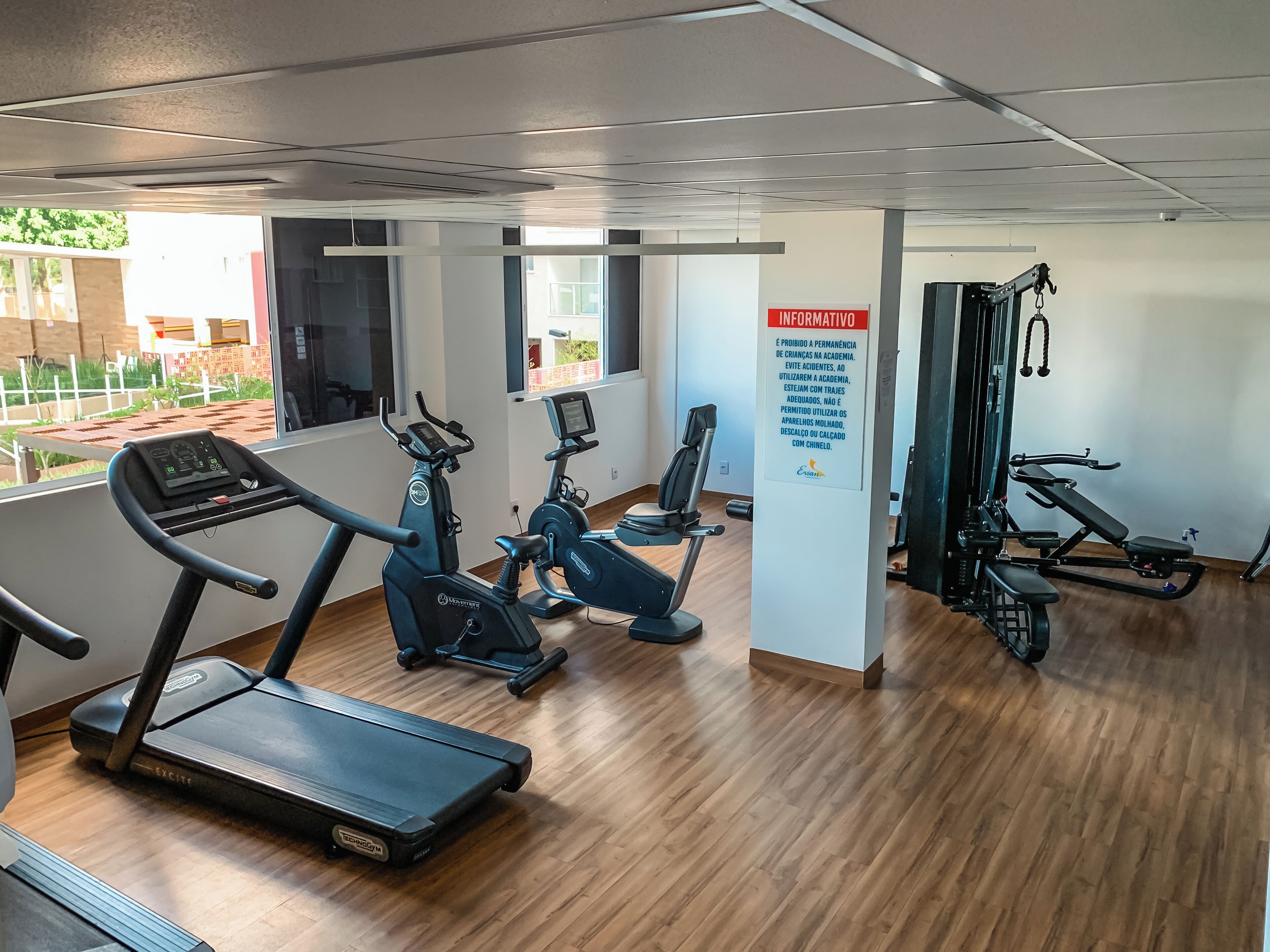 Academia com diversos equipamentos de exercício, incluindo esteiras e bicicletas ergométricas, e um aviso com instruções de segurança.