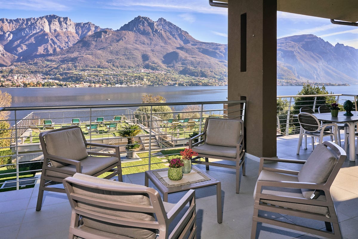 Um terraço com mesas ao ar livre, vista para um lago e montanhas ao fundo.