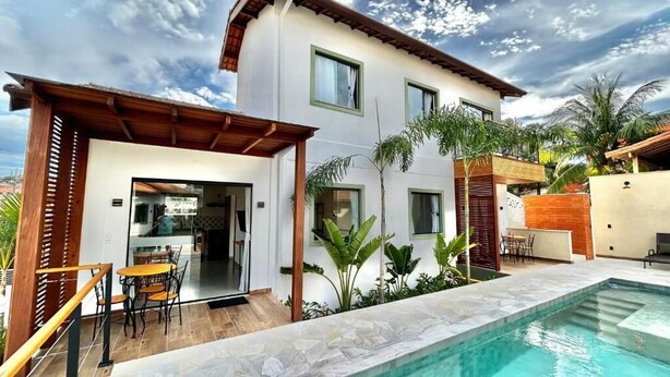 Uma casa moderna de dois andares com piscina, um pátio com mobiliário externo e um jardim com plantas tropicais.