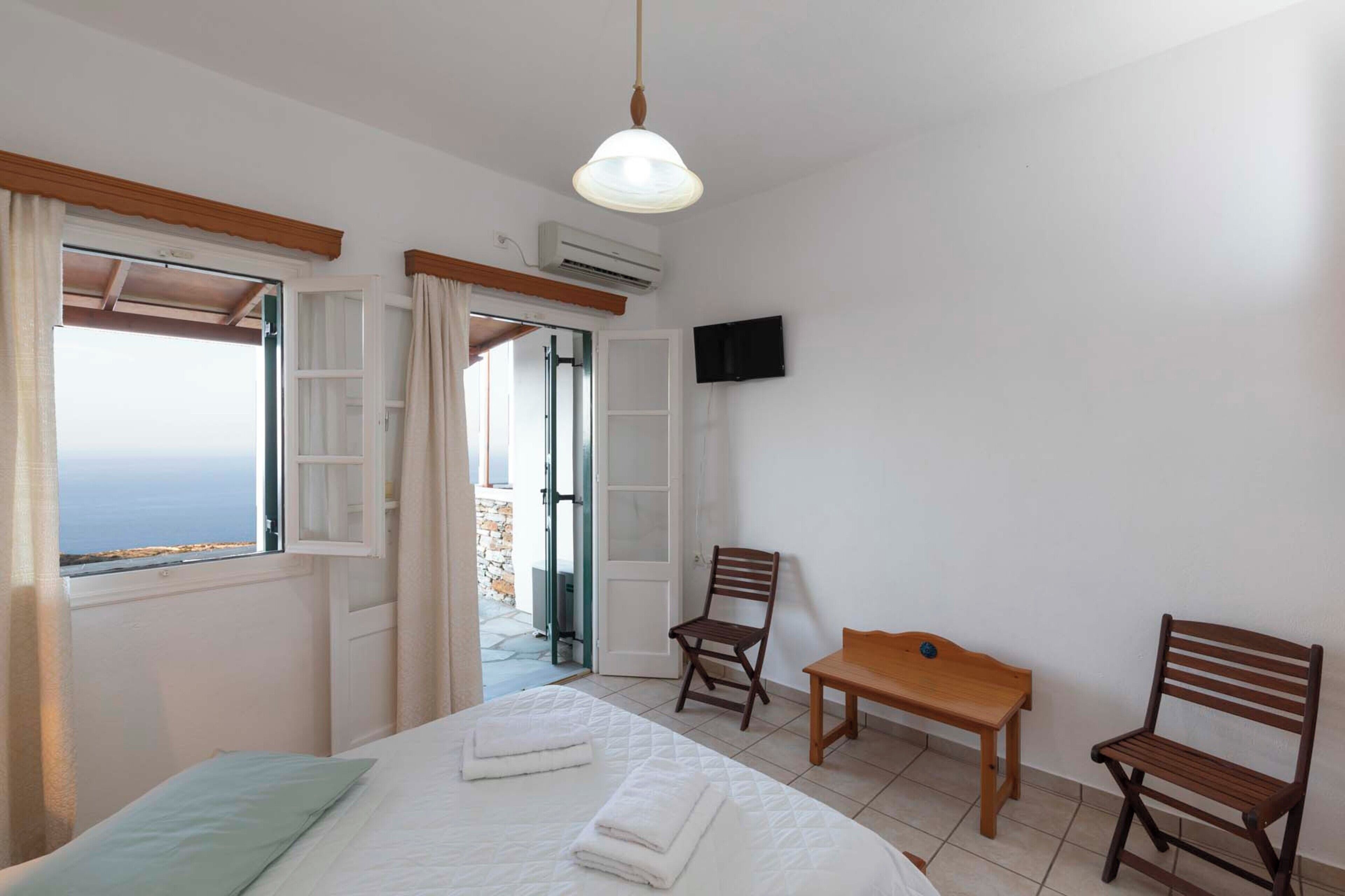 Quarto com cama, duas cadeiras de madeira, uma mesa de madeira, TV fixada na parede e vista através de uma porta aberta.