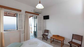 Quarto com cama, duas cadeiras de madeira, uma mesa de madeira, TV fixada na parede e vista através de uma porta aberta.