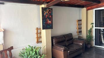 Uma área de estar coberta ao ar livre com um banco de madeira, um sofá de couro marrom e um vaso com planta.