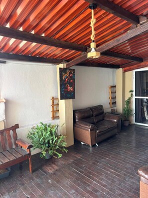 Uma área de estar coberta ao ar livre com um banco de madeira, um sofá de couro marrom e um vaso com planta.