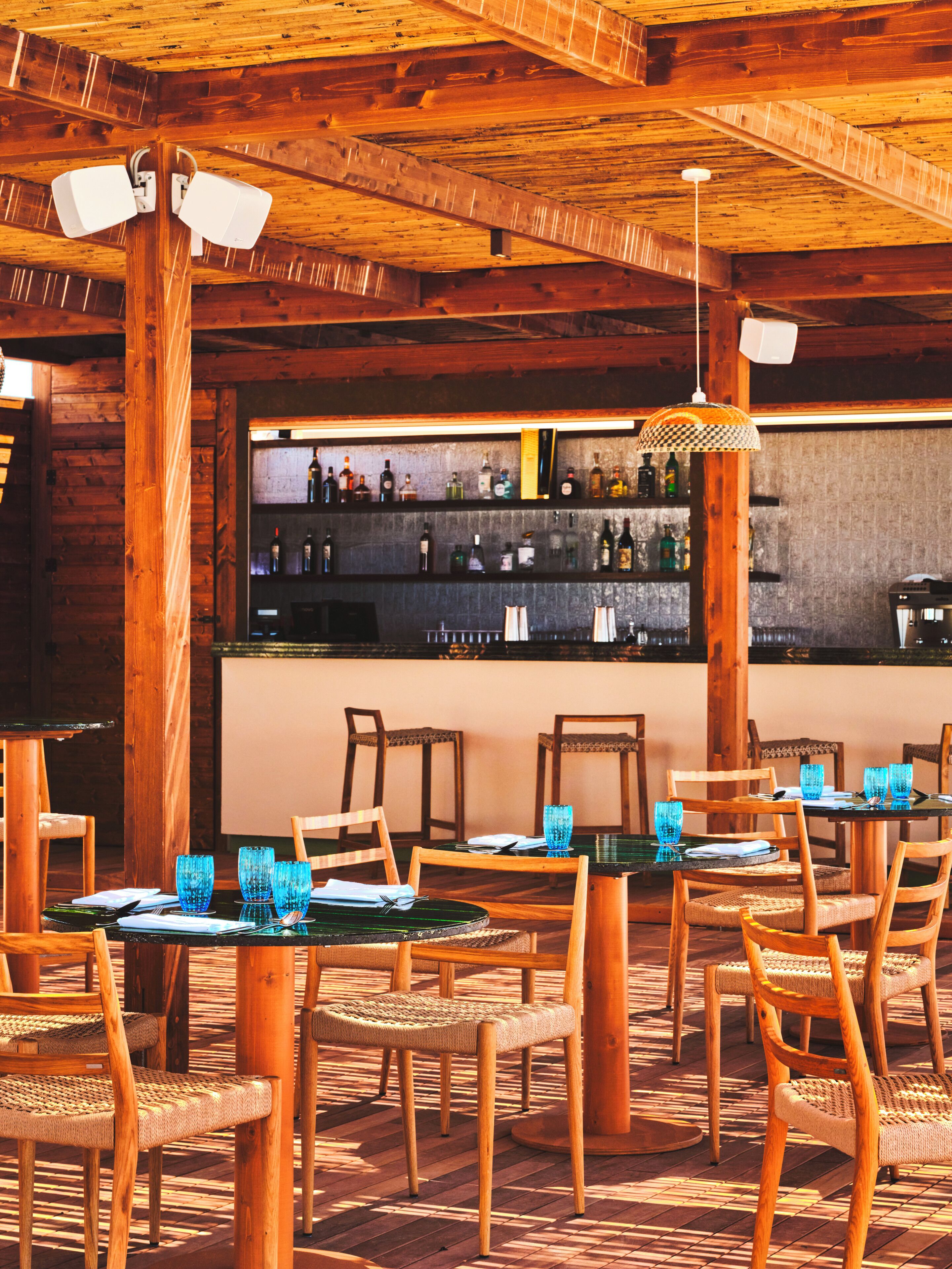 Restaurante com mobiliário de madeira, mesas com copos de vidro azul e um bar com prateleiras cheias de garrafas.