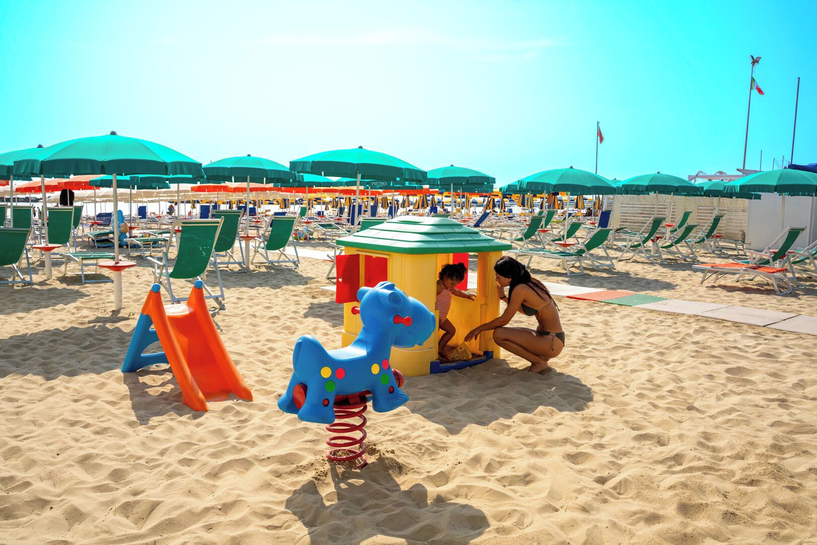Uma praia com guarda-sóis verdes, espreguiçadeiras e uma área de recreação com um escorrega e um cavalo de brinquedo azul.