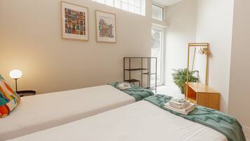 Quarto de hotel com duas camas, uma mesinha de cabeceira, luminária, espelho e quadros nas paredes.
