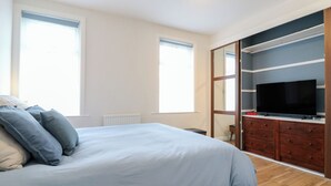 Quarto com uma cama, uma televisão sobre um móvel de madeira e duas janelas.