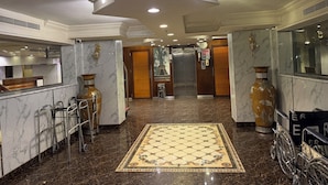 Um saguão de hotel com piso de mármore, um tapete decorativo central e um elevador, além de uma cadeira de rodas.