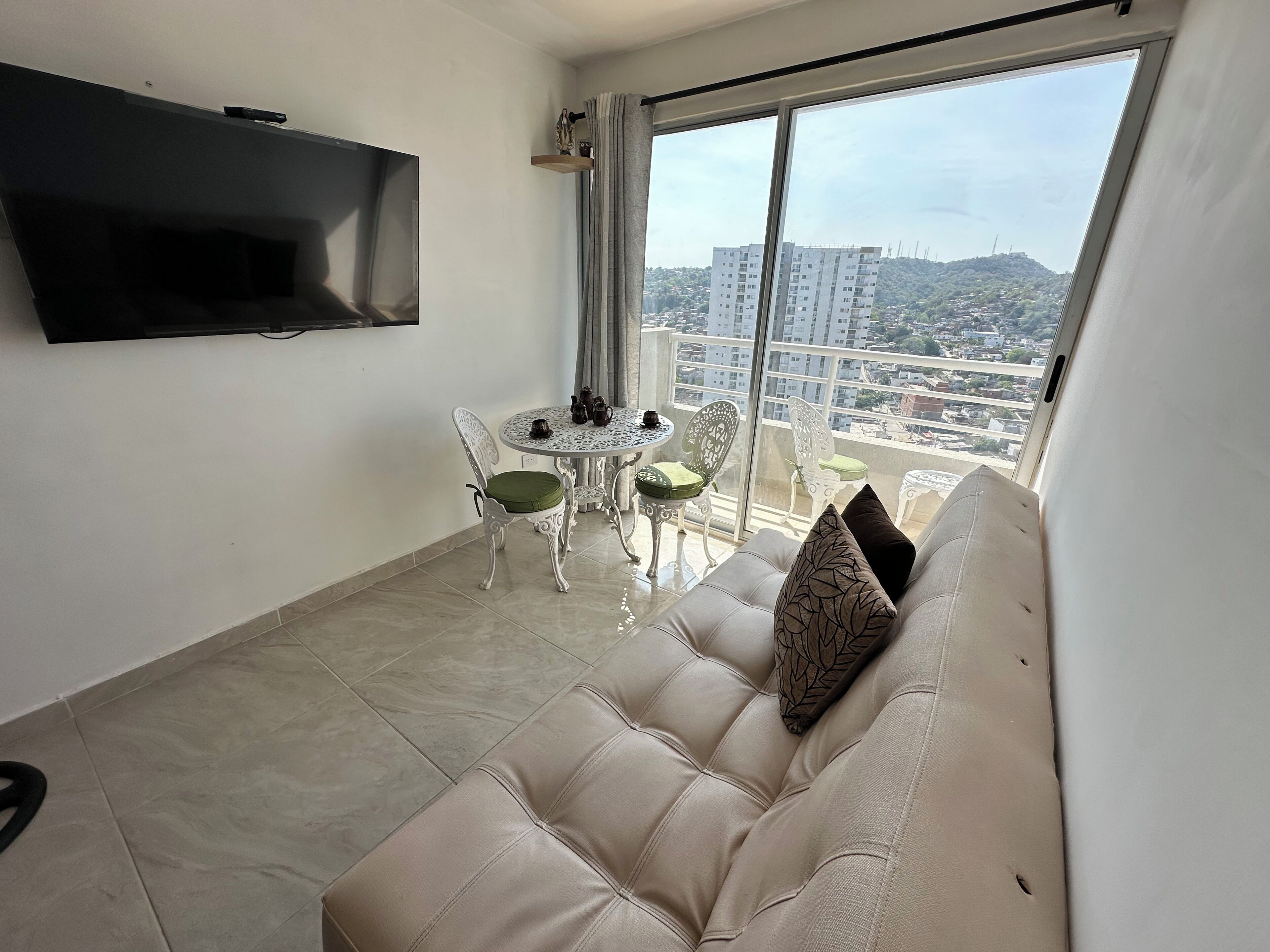 Uma sala de estar moderna com um sofá bege, uma mesinha redonda e duas cadeiras brancas. Uma televisão de tela plana está fixada na parede, e uma grande janela oferece vista para o horizonte urbano.