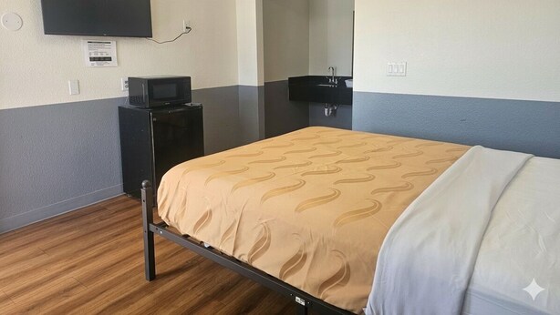 Quarto de hotel com uma cama, televisão de tela plana na parede, micro-ondas, pia e uma porta que dá para outro cômodo.