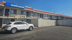 Um motel com um letreiro de 'bem-vindo', um carro branco estacionado na frente e uma área de estacionamento cercada.