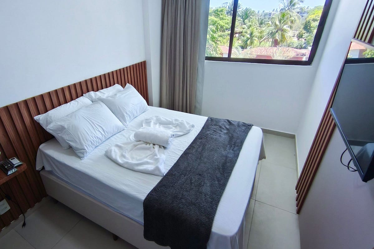 Cama bem arrumada com lençóis e toalhas brancas, cabeceira de madeira, mesa de cabeceira com abajur e janela com vista para o verde.