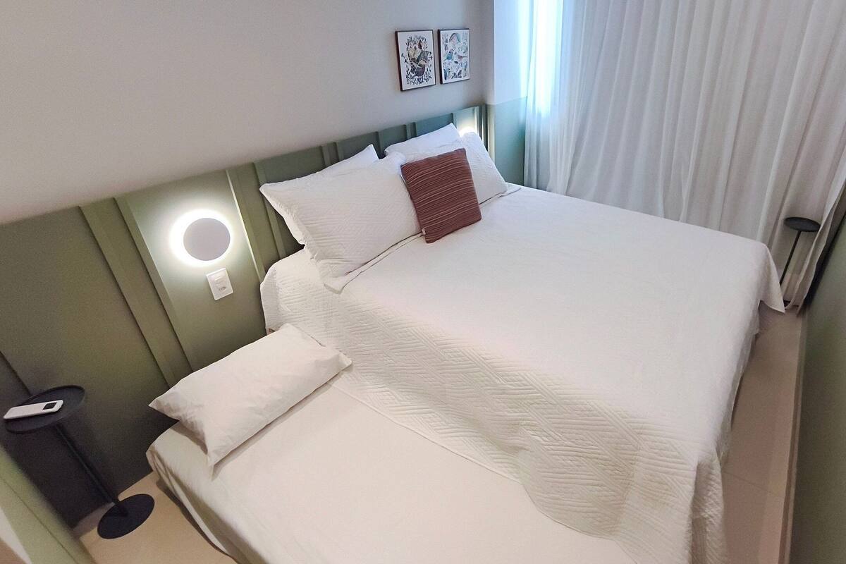 Cama bem arrumada com roupa de cama branca e travesseiro marrom, mesa de cabeceira com telefone e luminária fixada na parede.
