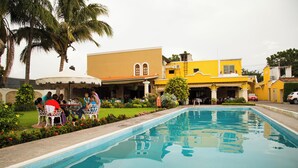 Uma piscina com borda azulejada, rodeada por um jardim com palmeiras e um edifício com fachada amarela.