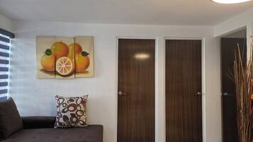 Um quarto com um sofá marrom escuro, uma almofada estampada, um quadro de frutas na parede e duas portas de madeira fechadas.
