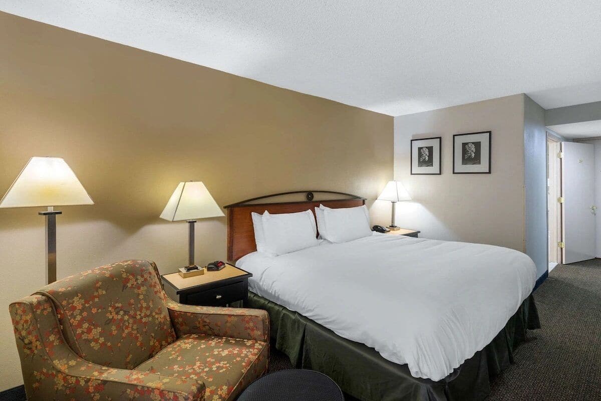 Quarto de hotel com cama, duas luminárias de cabeceira, uma poltrona estampada e uma mesinha de cabeceira.