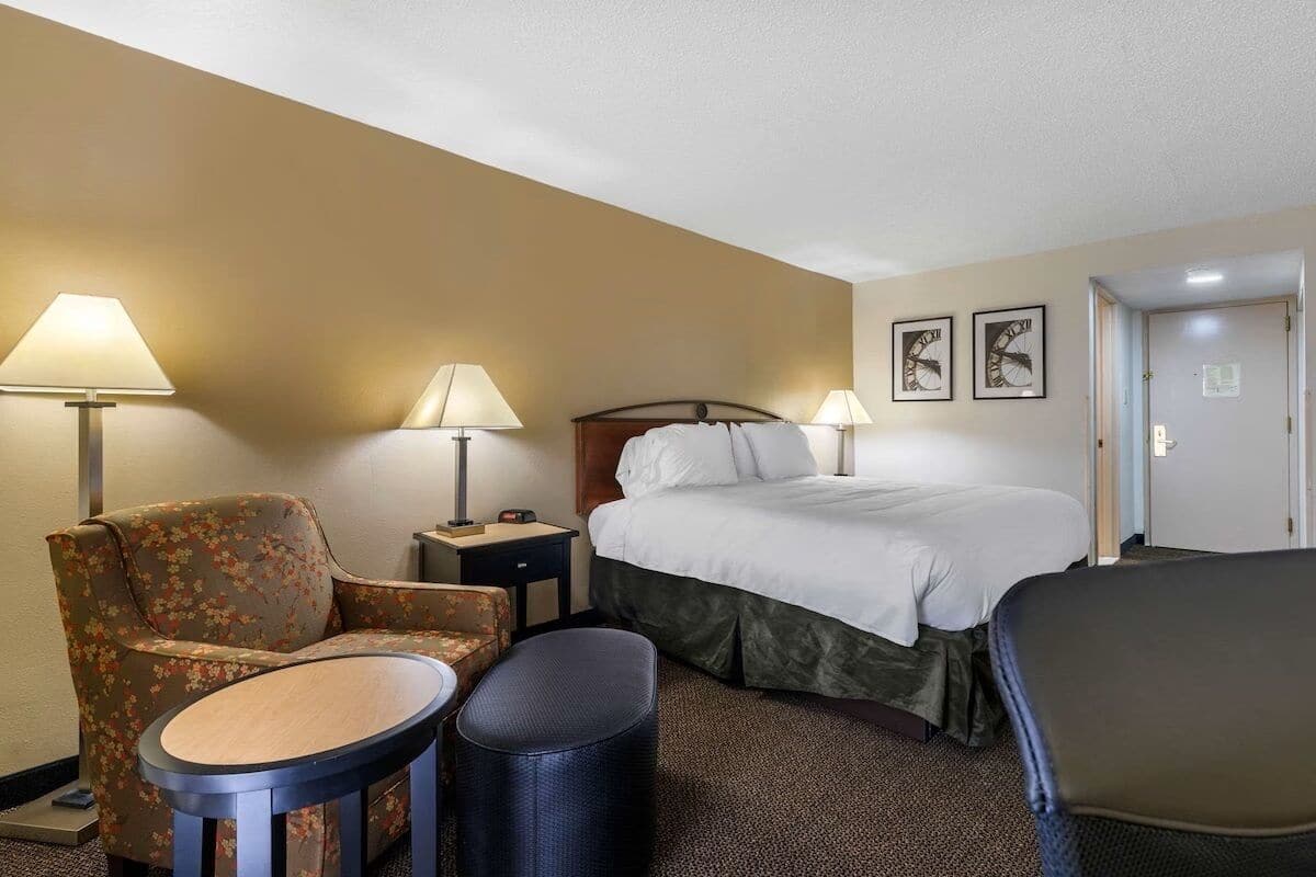 Quarto de hotel com uma cama, duas cadeiras, uma mesinha, duas luminárias de cabeceira e dois relógios de parede.